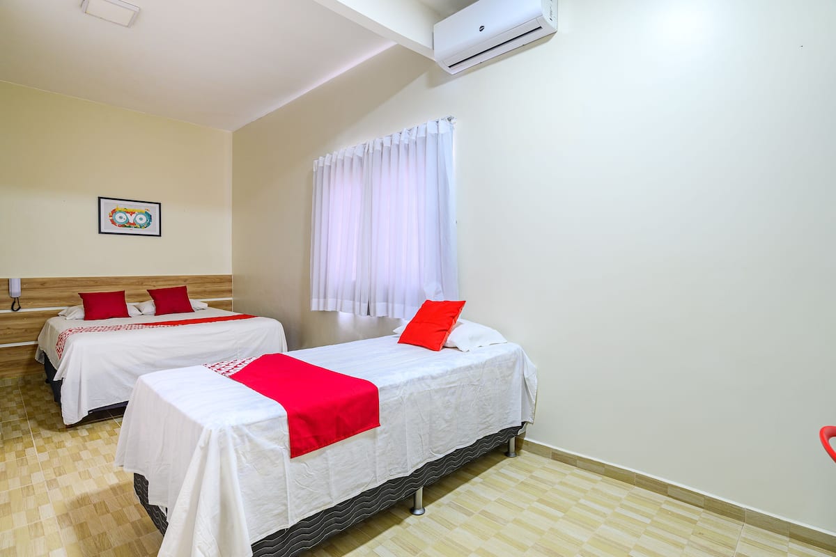 Quarto de hotel com duas camas, cada uma com roupa de cama branca e detalhes vermelhos, uma janela com cortinas brancas e um ar condicionado fixado na parede.