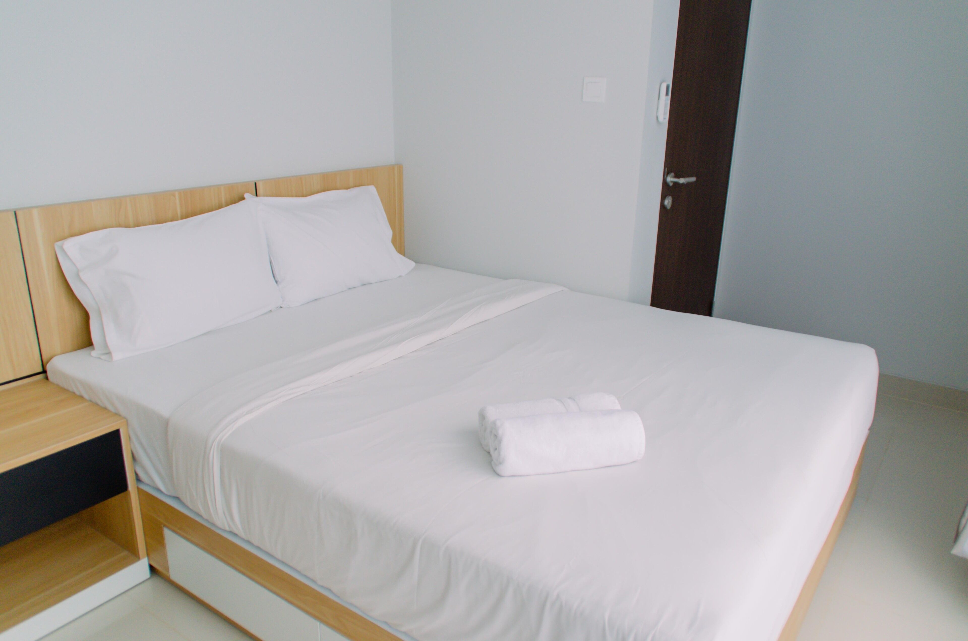 Cama bem arrumada com lençóis brancos e toalha dobrada, cabeceira de madeira e mesa de cabeceira de madeira com gaveta.