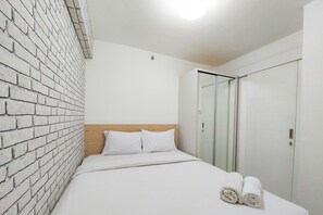 Quarto de hotel com parede de tijolos aparentes, cabeceira de madeira, cama com roupa de cama branca e armário de vidro com uma toalha em cima.