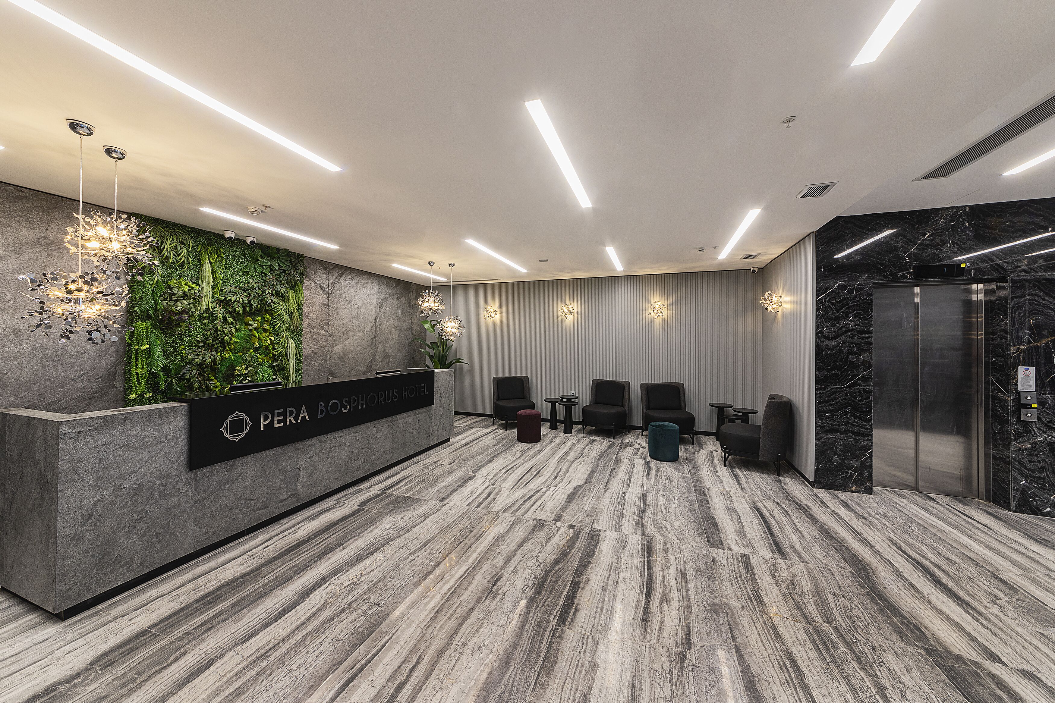 Uma área de recepção moderna de hotel, com balcão de atendimento, espaço de estar e parede verde com plantas.