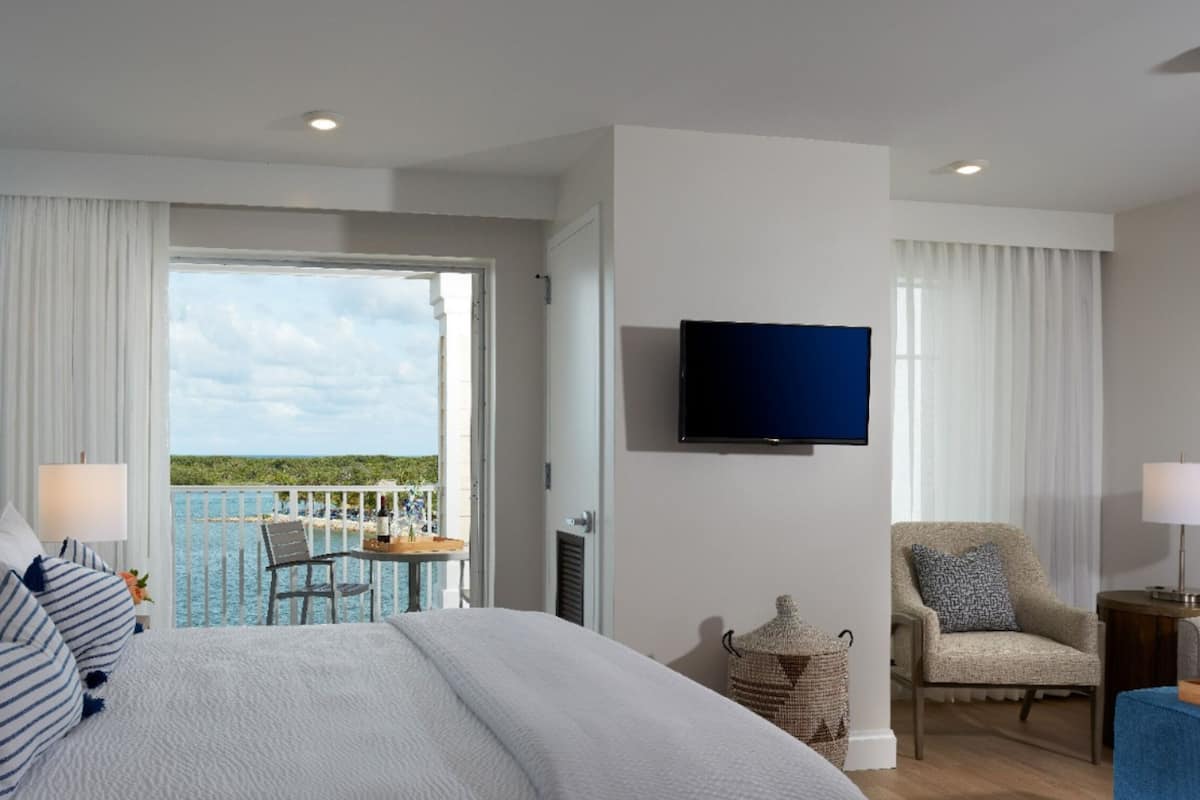 Quarto de hotel com uma cama grande, televisão, varanda com vista para o mar e uma cadeira.