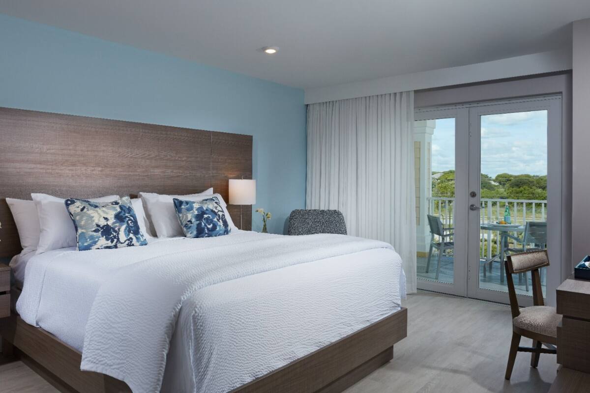 Quarto de hotel moderno com uma cama grande, duas luminárias de cabeceira, uma mesa de cabeceira, uma cadeira, uma escrivaninha e uma varanda com vista.
