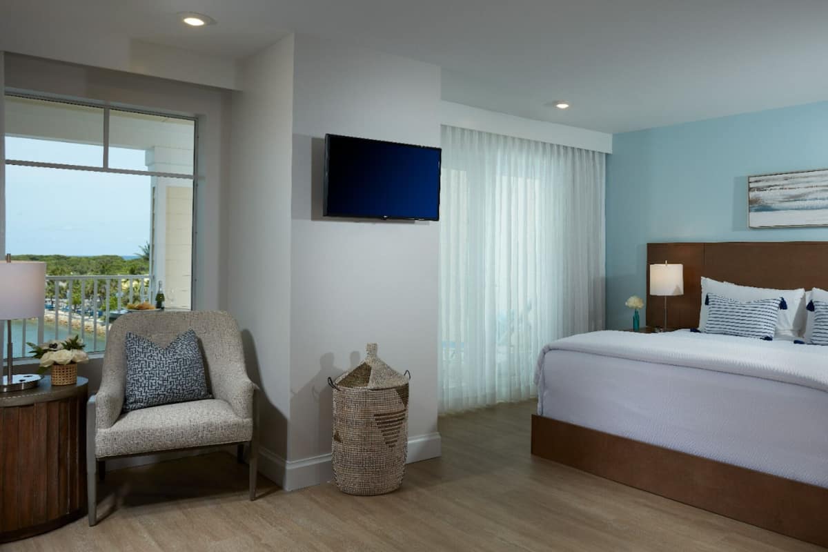Quarto de hotel com cama, poltrona, televisão, janela com cortinas e uma obra de arte na parede.