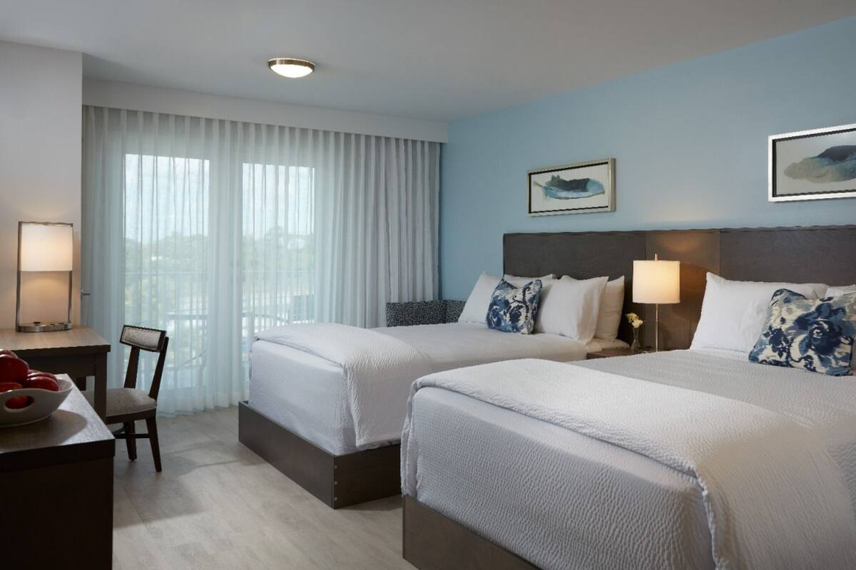 Quarto de hotel com duas camas, uma mesa, uma cadeira e uma janela com cortinas.