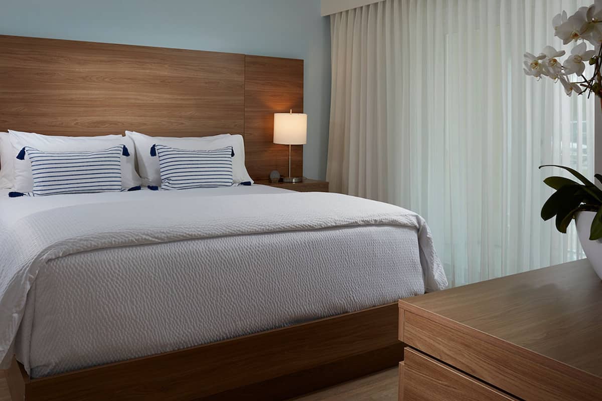 Cama bem arrumada com roupa de cama branca e almofadas listradas, com duas luminárias de cabeceira em mesas de cabeceira de madeira.
