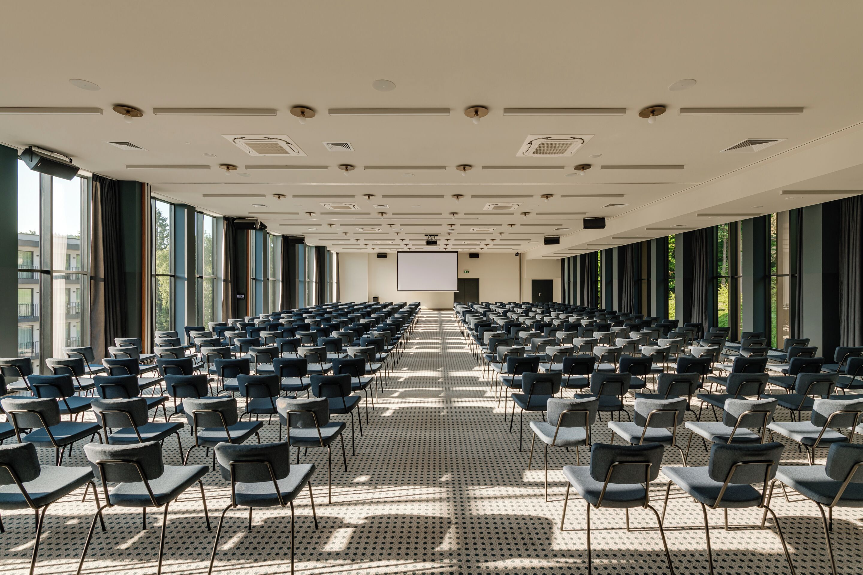 Sala de conferências com fileiras de cadeiras voltadas para uma tela de projeção, amplas janelas em um dos lados e piso com padrão.