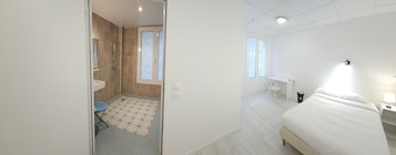 Quarto com cama, escrivaninha e cadeira. Há uma área de chuveiro com piso cerâmico e um banquinho.