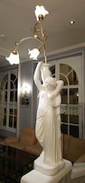 Uma estátua branca com manto, um lustre dourado decorativo e uma mesa de vidro num espaço com sofá bege e janelas arqueadas.