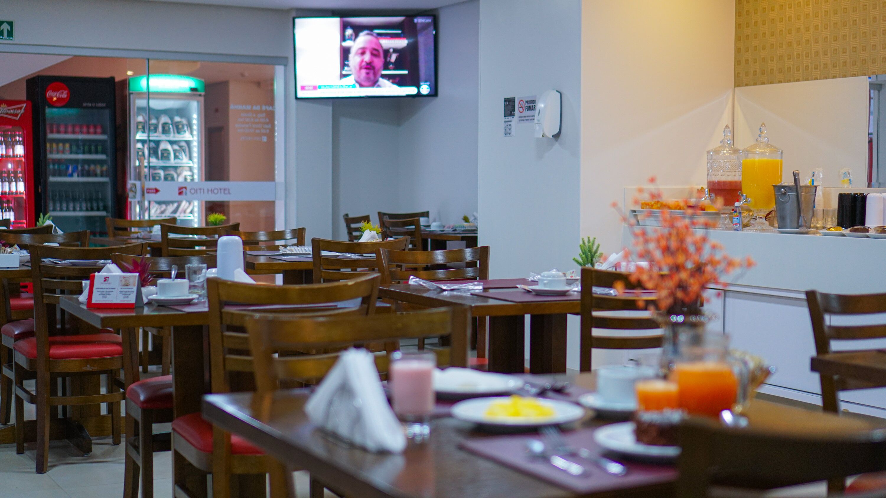 Uma sala de jantar de hotel com mesas e cadeiras de madeira, uma televisão na parede e um balcão de café da manhã com suco de laranja e outras bebidas.