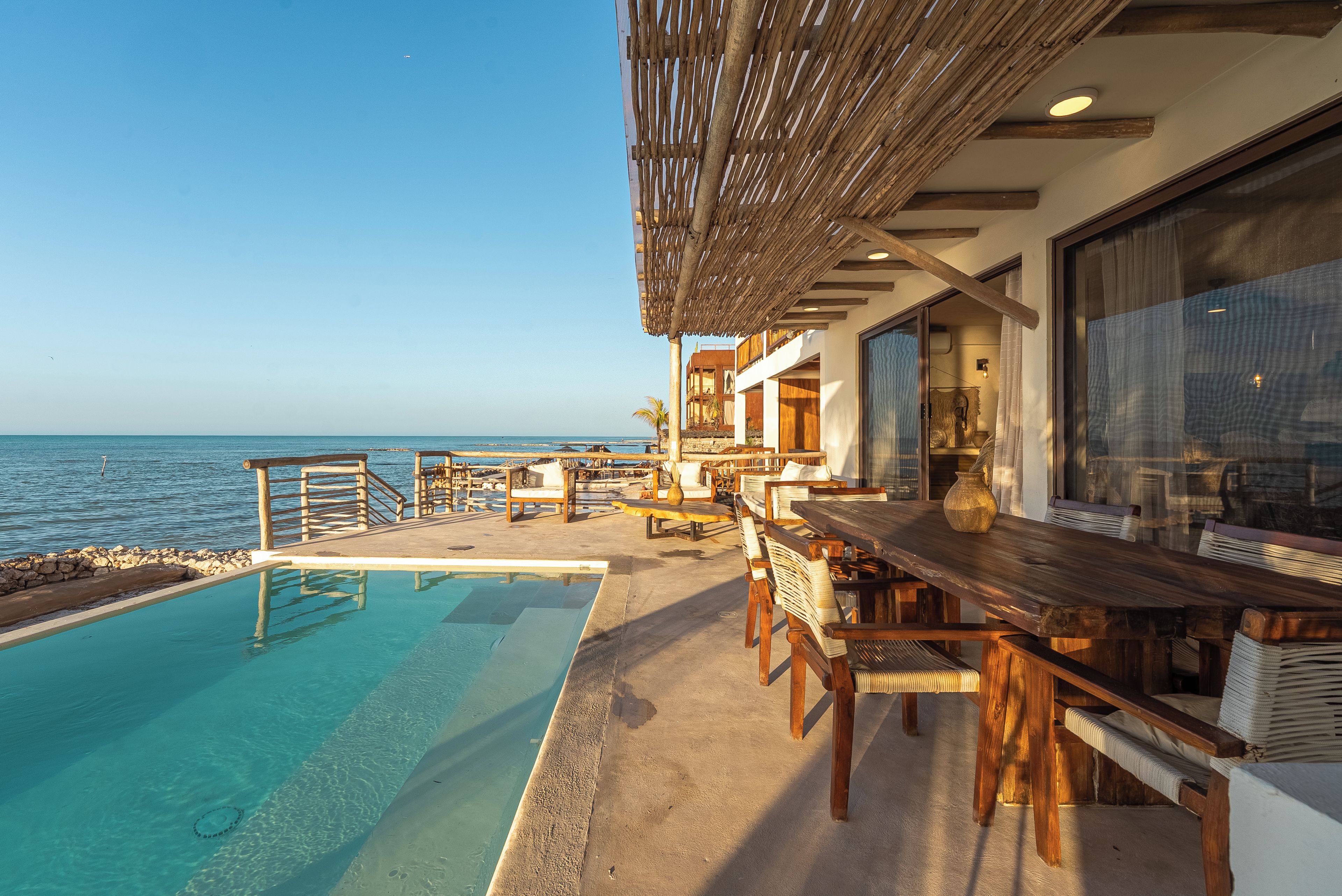 Área da piscina com vista para o mar, mobiliário de madeira e um espaço coberto para sentar.