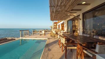 Área da piscina com vista para o mar, mobiliário de madeira e um espaço coberto para sentar.