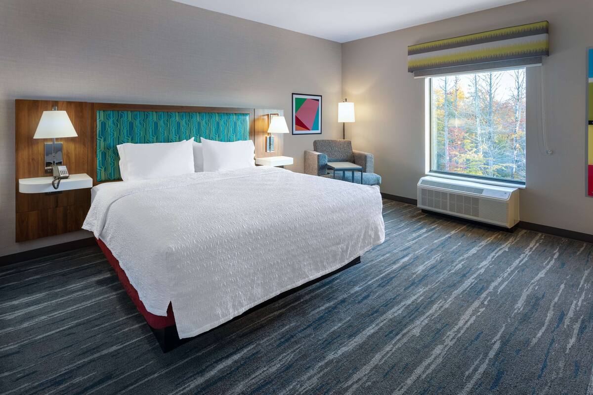 Quarto de hotel com uma cama grande, uma escrivaninha, uma cadeira, uma janela com vista para árvores e ar condicionado fixado na parede.