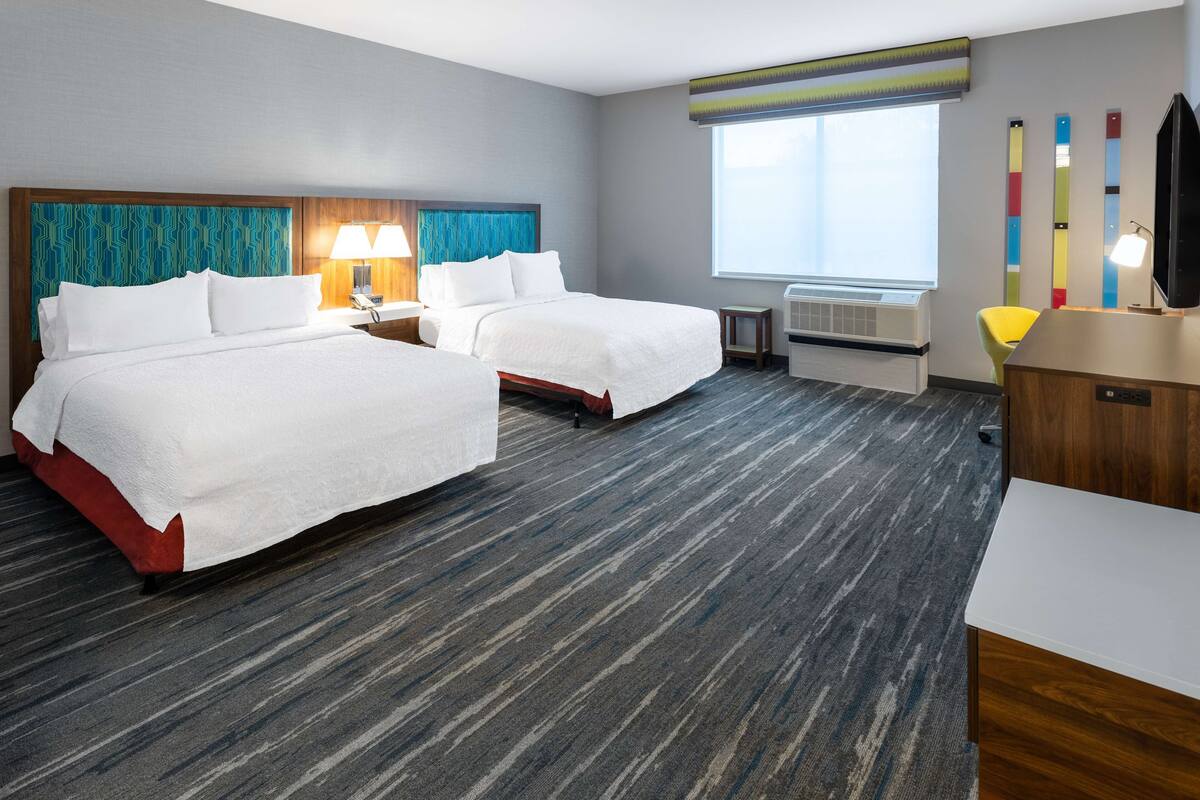 Quarto de hotel com duas camas, uma escrivaninha, uma cadeira, uma televisão e uma janela com vista.