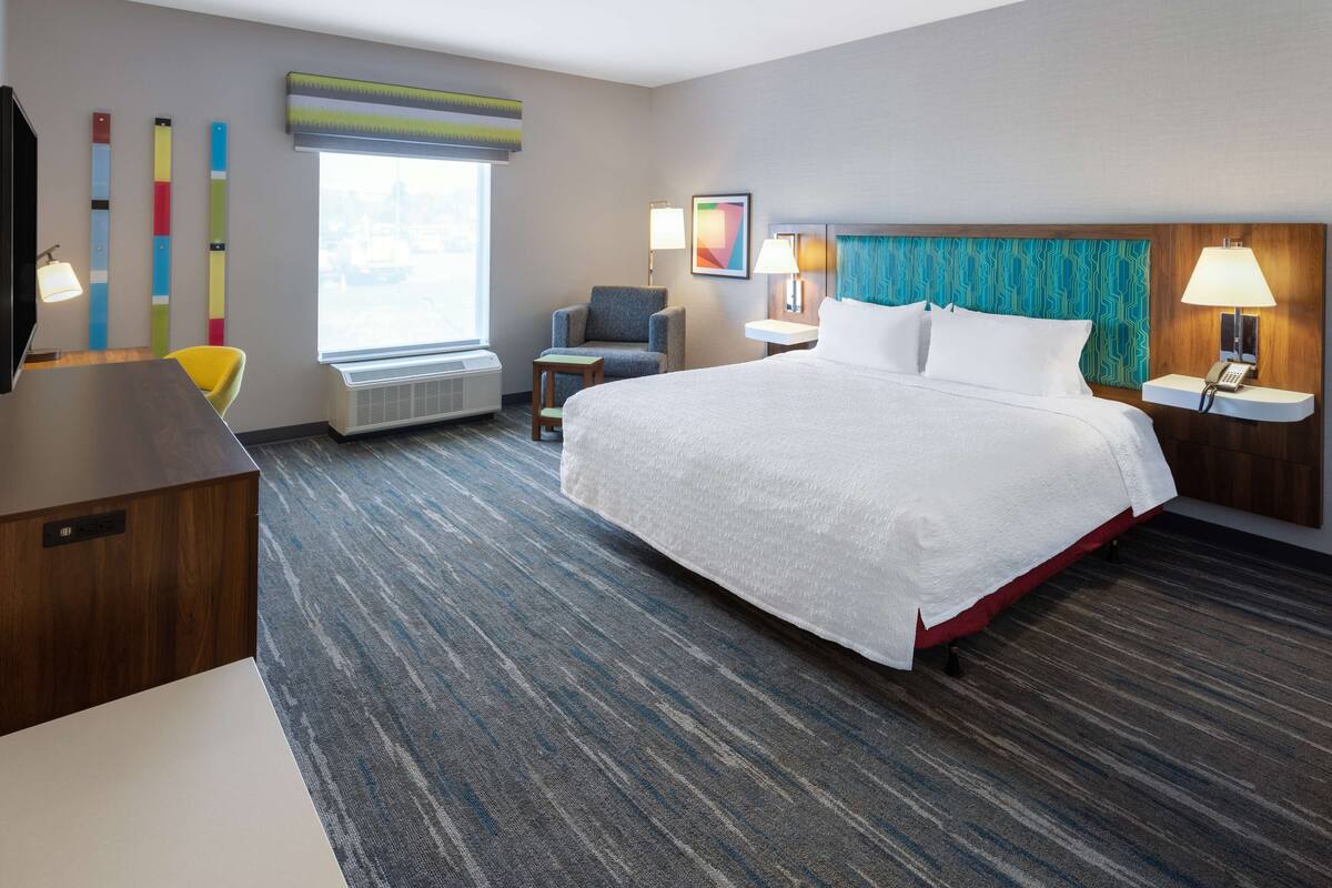 Quarto de hotel com uma cama grande, uma escrivaninha, uma cadeira, uma mesinha, um abajur e uma janela com persianas.