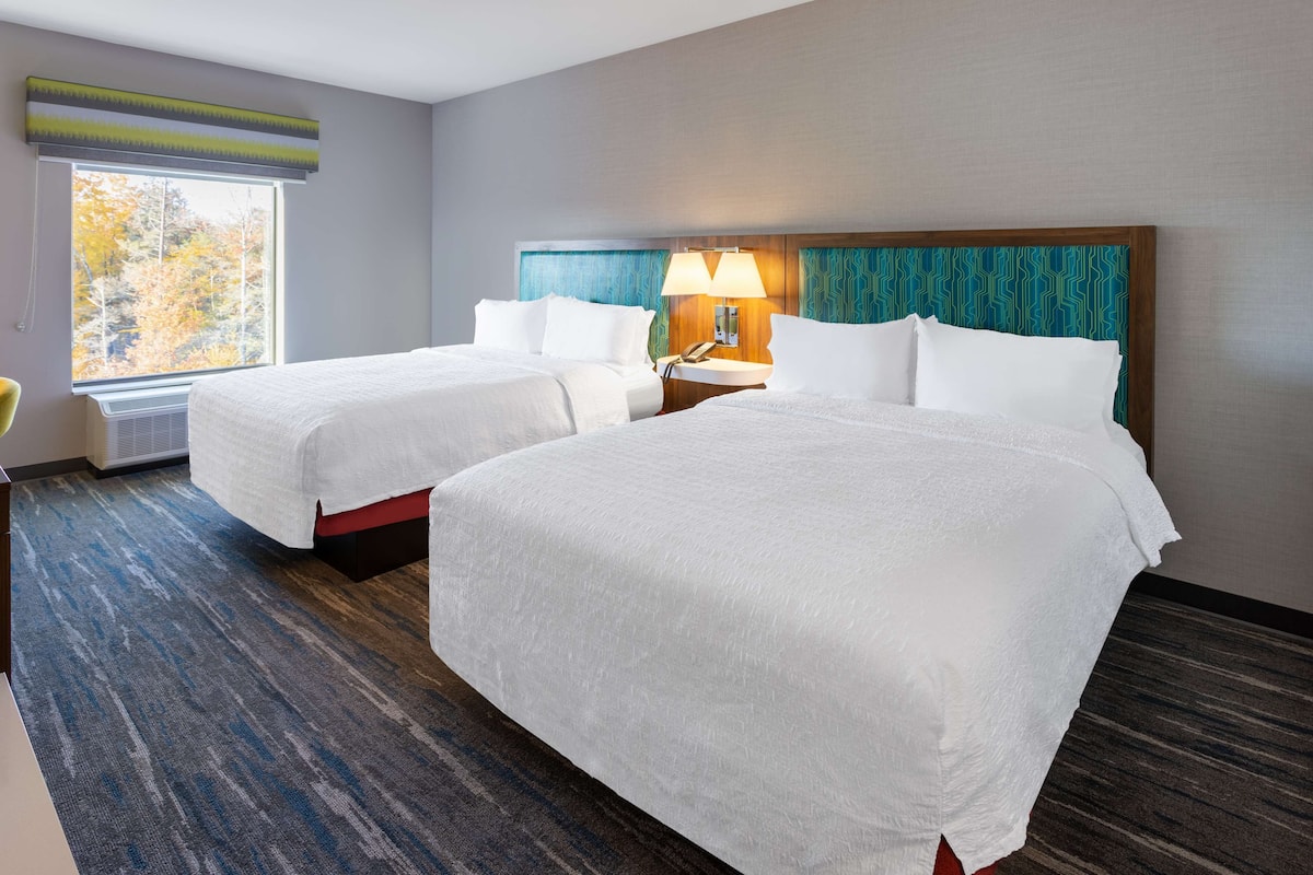 Quarto de hotel com duas camas, uma janela grande, luminária fixada na parede e cabeceira estampada.