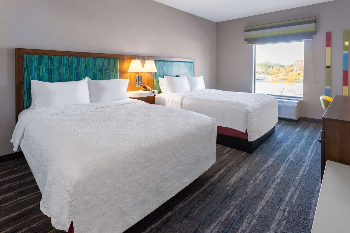Quarto de hotel com duas camas, mesa de cabeceira com abajur, janela com vista e piso coberto de carpete.
