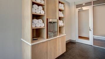 Quarto de hotel com estantes de madeira que contêm toalhas enroladas, um minibar e uma porta com painel de vidro jateado.