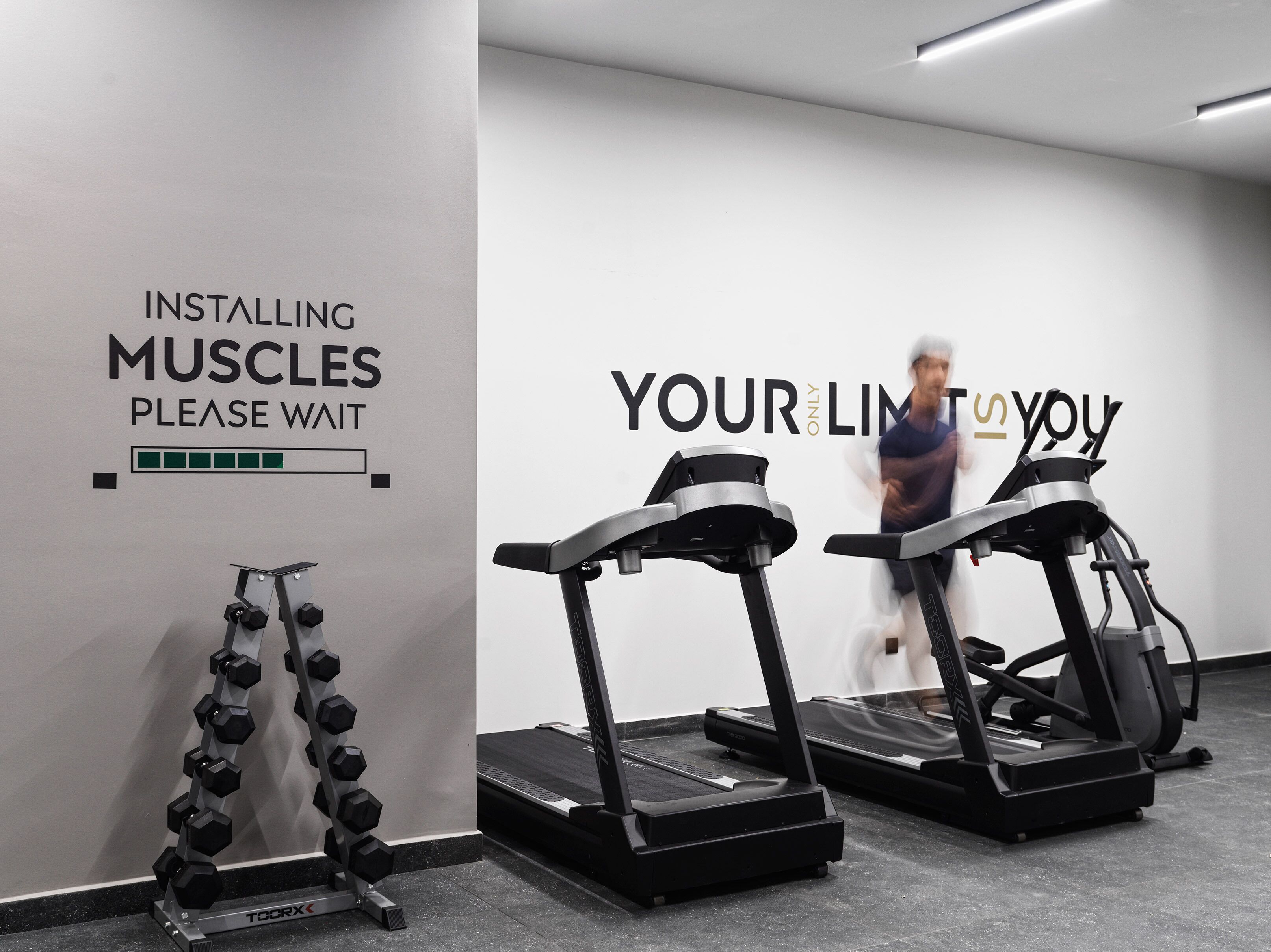 Uma academia com esteiras e halteres, uma placa com a mensagem 'FORTALECENDO MÚSCULOS, POR FAVOR, AGUARDE' e outra placa com a frase 'SUPERE SEUS LIMITES'.