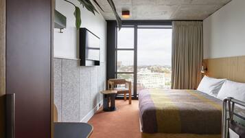 Quarto de hotel com uma janela grande, uma cama, uma cadeira e uma televisão fixada na parede.