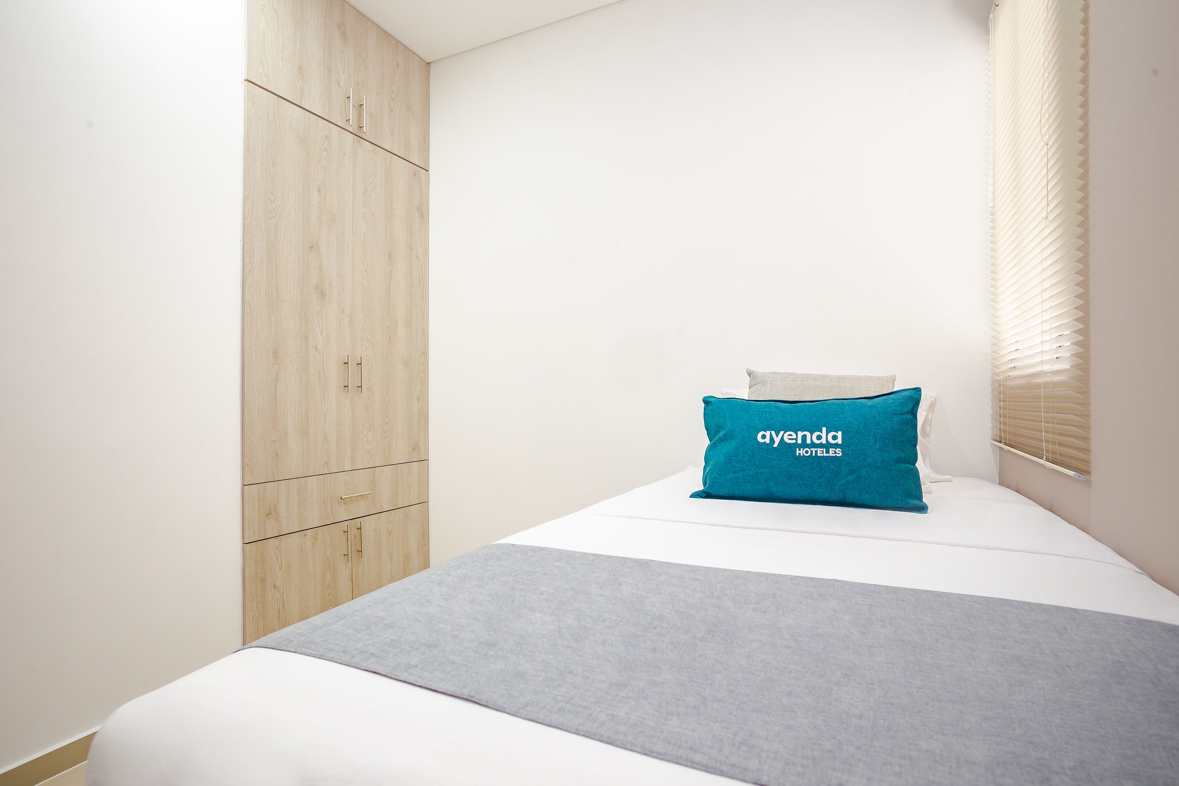 Quarto de hotel com cama, manta cinzenta, travesseiro azul-turquesa com a inscrição "ayenda hotels" e guarda-roupa de madeira. Inclui uma janela com persianas.