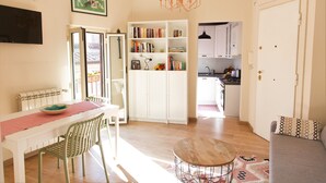 Uma sala de estar iluminada com uma mesa de jantar, cadeiras, uma estante e uma cozinha visível através de uma porta aberta.