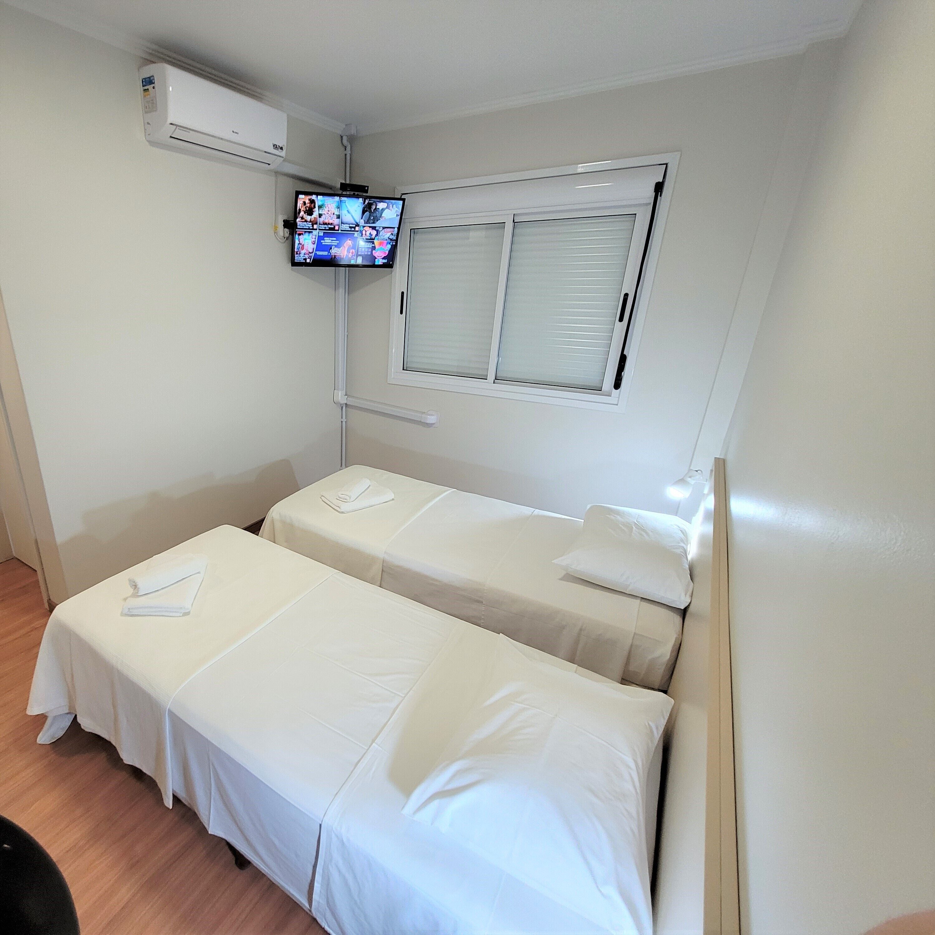 Quarto pequeno e limpo, com duas camas individuais, televisão na parede e janela com persianas.