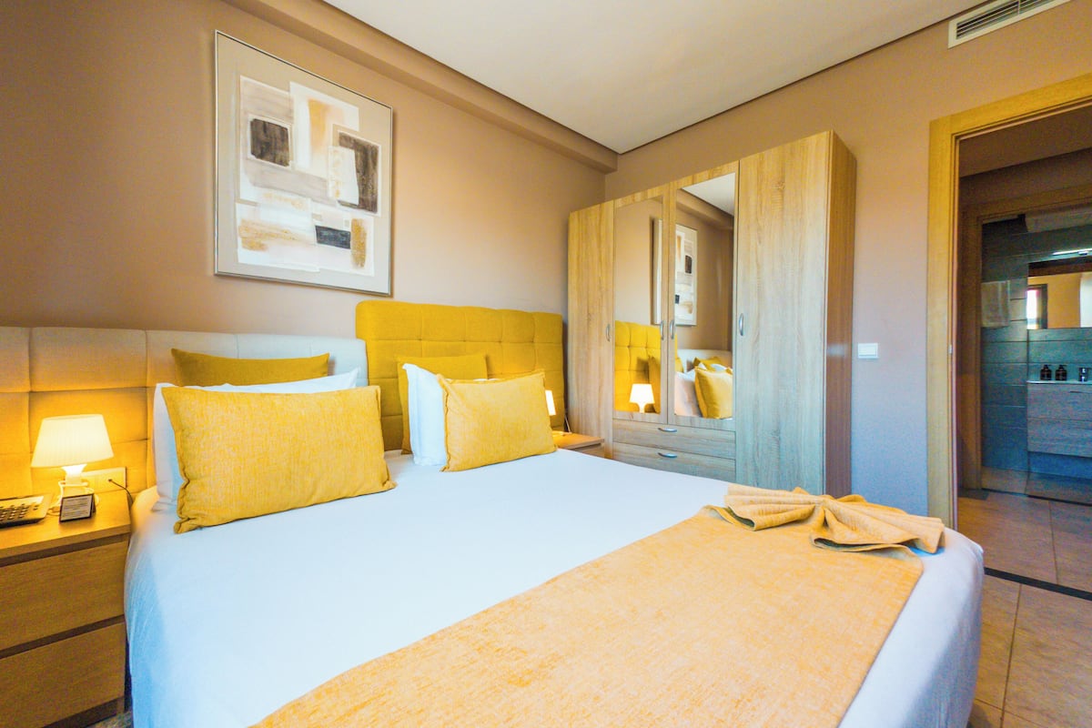 Quarto de hotel com uma cama grande, uma mesinha de cabeceira com abajur, telefone e espelho.