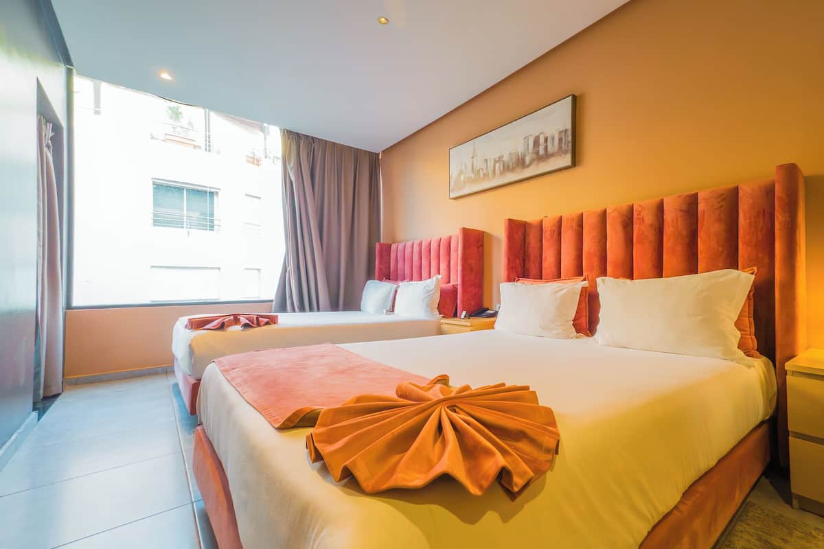 Quarto de hotel com duas camas, uma pintura de paisagem urbana, uma luminária e vista para prédios.
