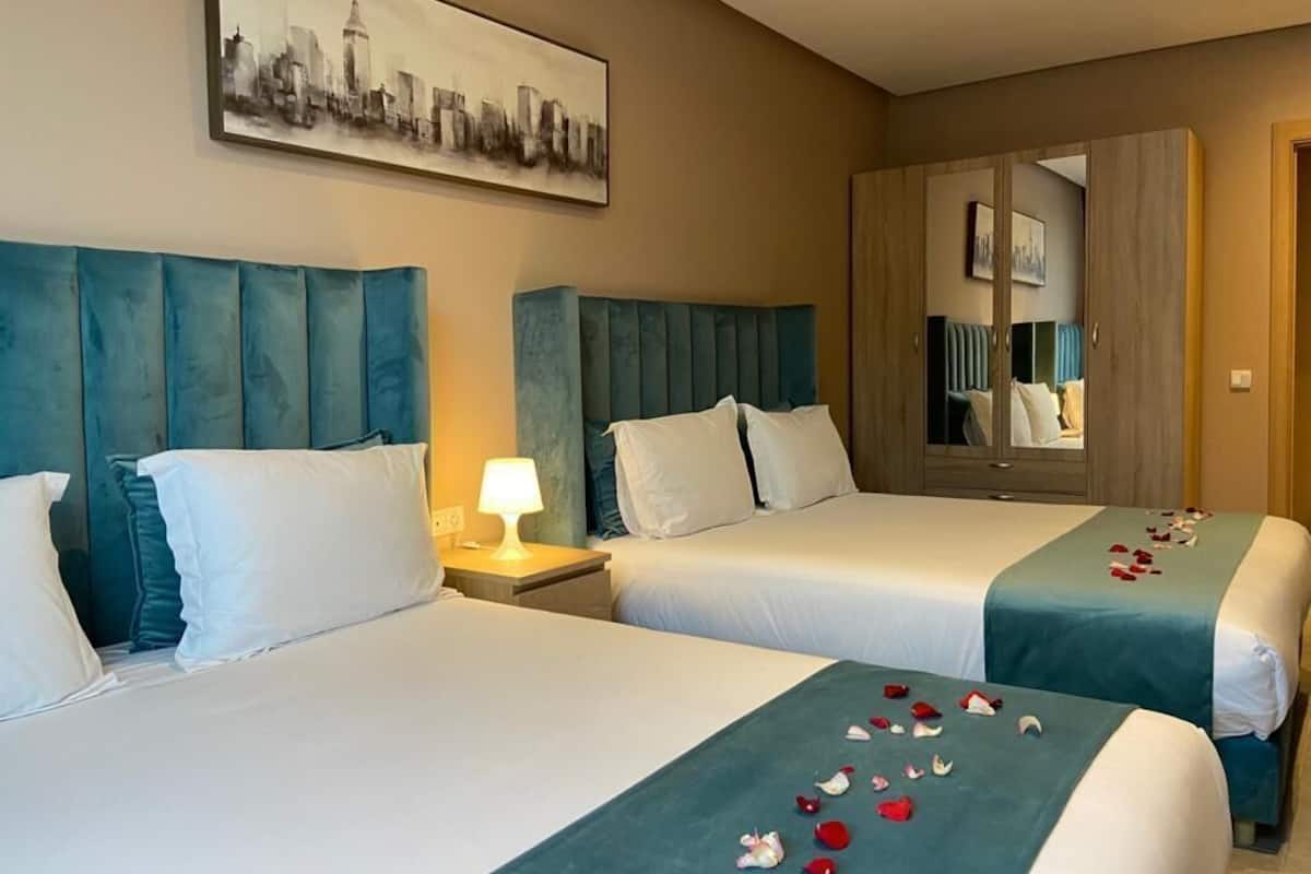 Quarto de hotel com duas camas, uma pintura de paisagem urbana, um espelho e um guarda-roupa de madeira.