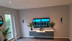 Uma sala de estar com televisão de tela plana na parede, piso de madeira e uma janela grande.