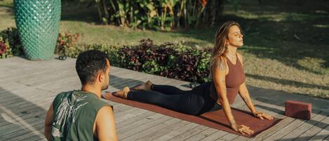 Duas pessoas praticando yoga em tapetes de treino ao ar livre, num jardim com um deck de madeira e vasos de plantas.