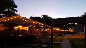 Uma área de restaurante ao ar livre, com luzes de corda, mesas de madeira e uma estrutura com telhado de palha.