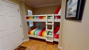 Uma cama de solteiro com roupa de cama colorida em um quarto com paredes bege, uma porta e uma janela com persianas.