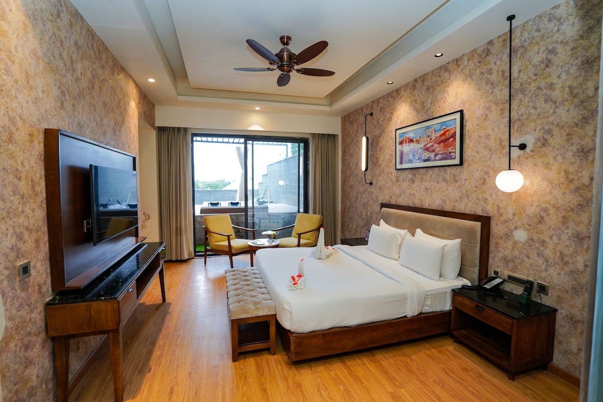 Quarto de hotel com cama, televisão, escrivaninha, cadeira, mesinha e ventilador de teto.