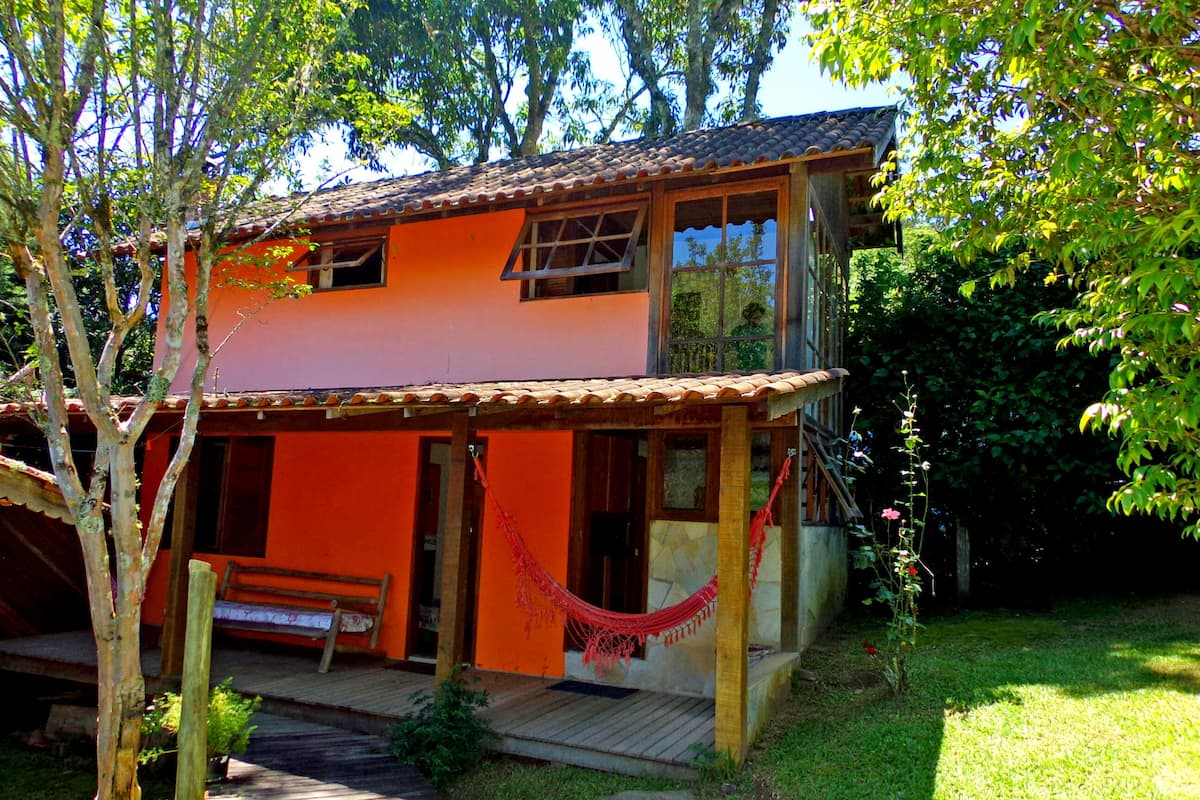 Uma casa de dois andares com fachada vermelha, varanda de madeira e uma rede.
