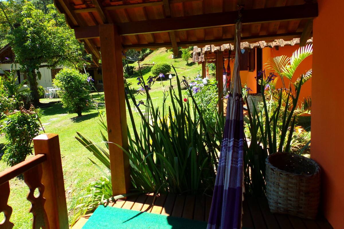 Um deck de madeira com uma rede, cercado por vegetação exuberante e vista para um jardim.