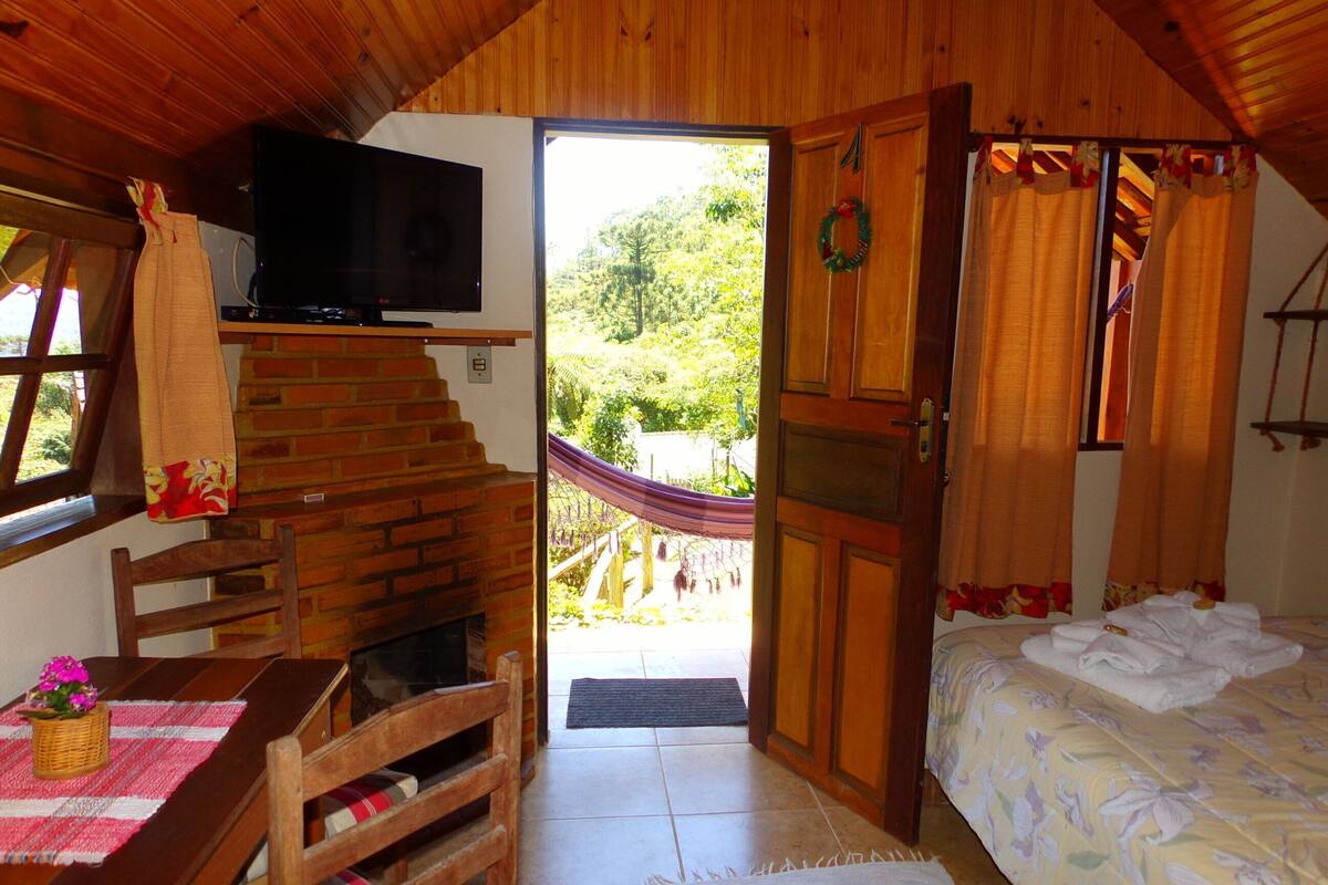 Um quarto acolhedor com televisão, uma cama, uma cadeira de madeira, uma mesa com um cesto de flores e vista para um jardim.