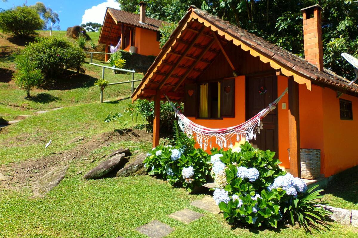 Uma pequena casa térrea com telhado vermelho e paredes alaranjadas, rodeada por vegetação exuberante e uma rede.