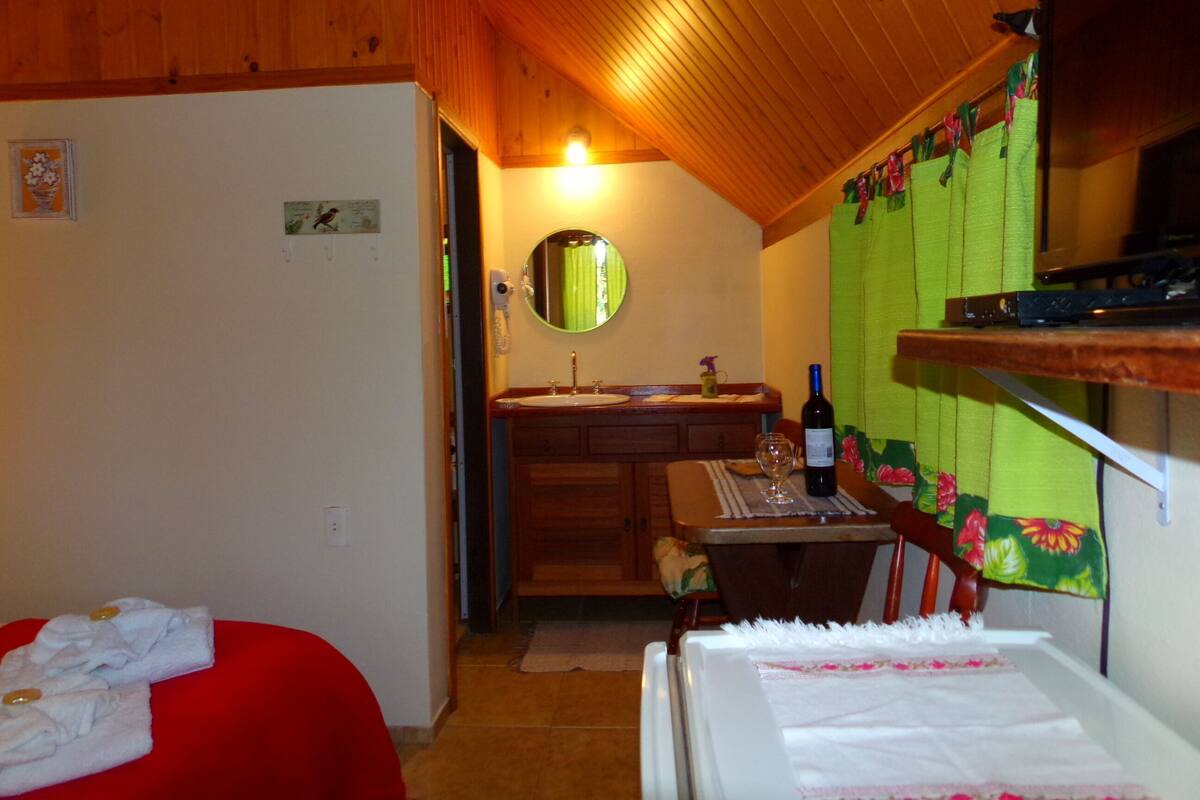 Quarto com teto de madeira, uma cama, uma mesa com uma garrafa de vinho, um espelho e cortina verde.