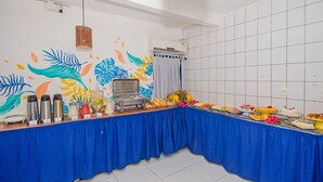 Disposição de buffet com diversos alimentos e bebidas, um mural decorativo e uma parede revestida de azulejos.