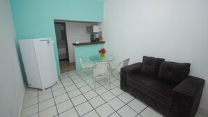 Um espaço compacto com cozinha americana, área de jantar com mesa de vidro e cadeiras brancas, e um sofá marrom escuro.