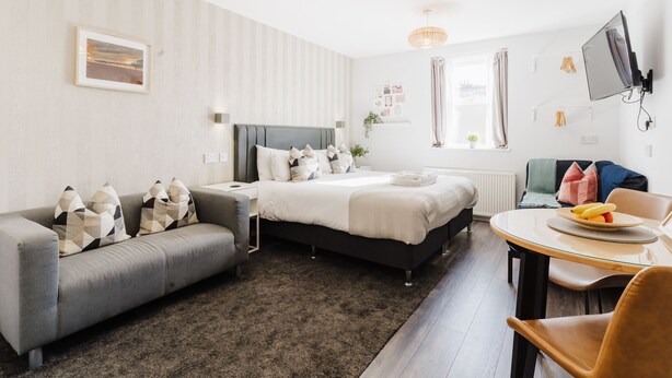 Quarto de hotel moderno com cama, sofá, mesa de jantar com cadeiras e televisão na parede.