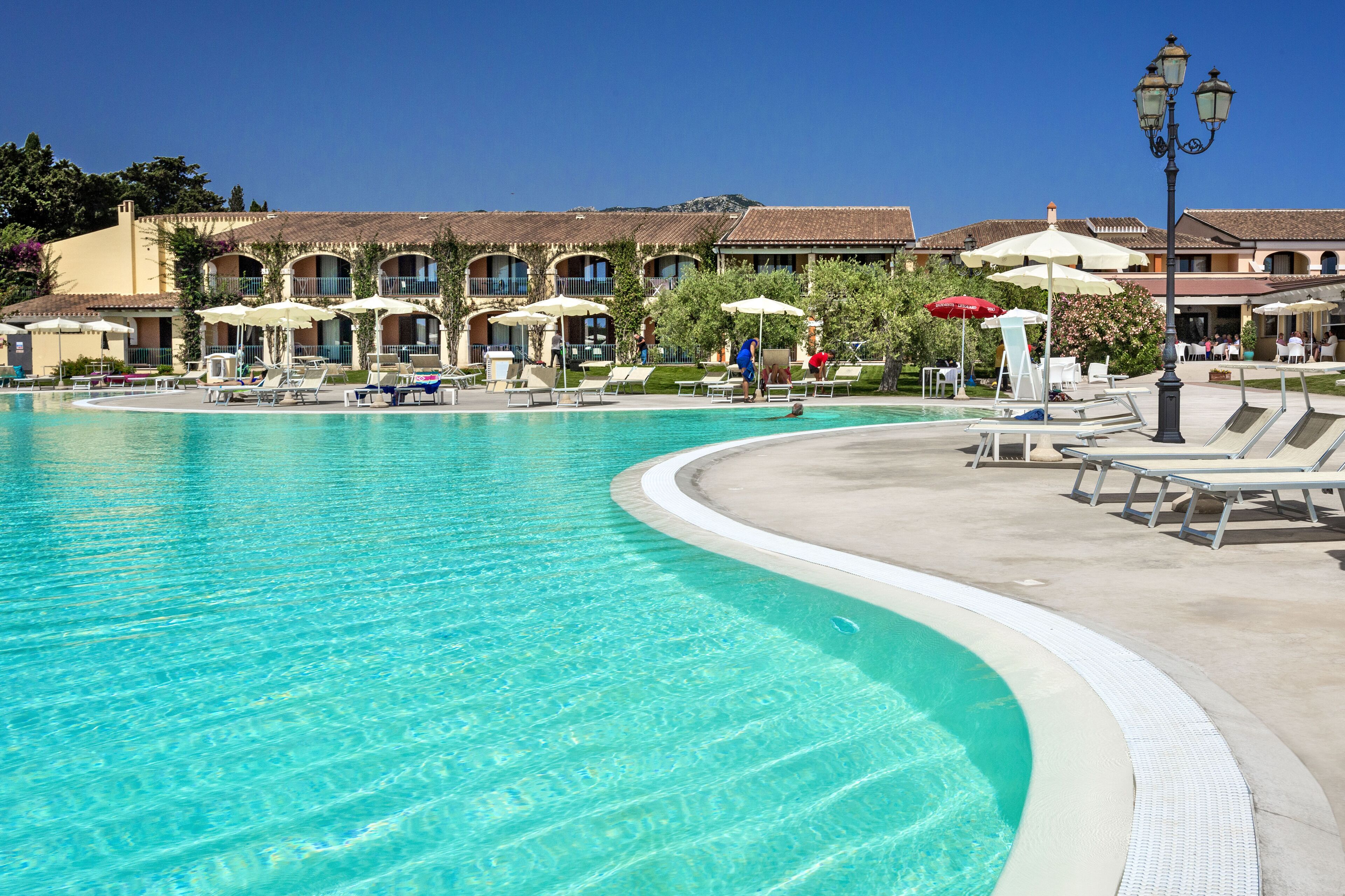 Piscina com espreguiçadeiras e guarda-sóis, rodeada por edifícios com varandas e céu claro.