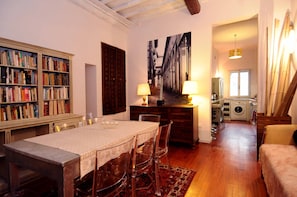 Uma sala de jantar com uma mesa posta para quatro pessoas, uma estante de livros, um quadro e uma área de serviço ao fundo.