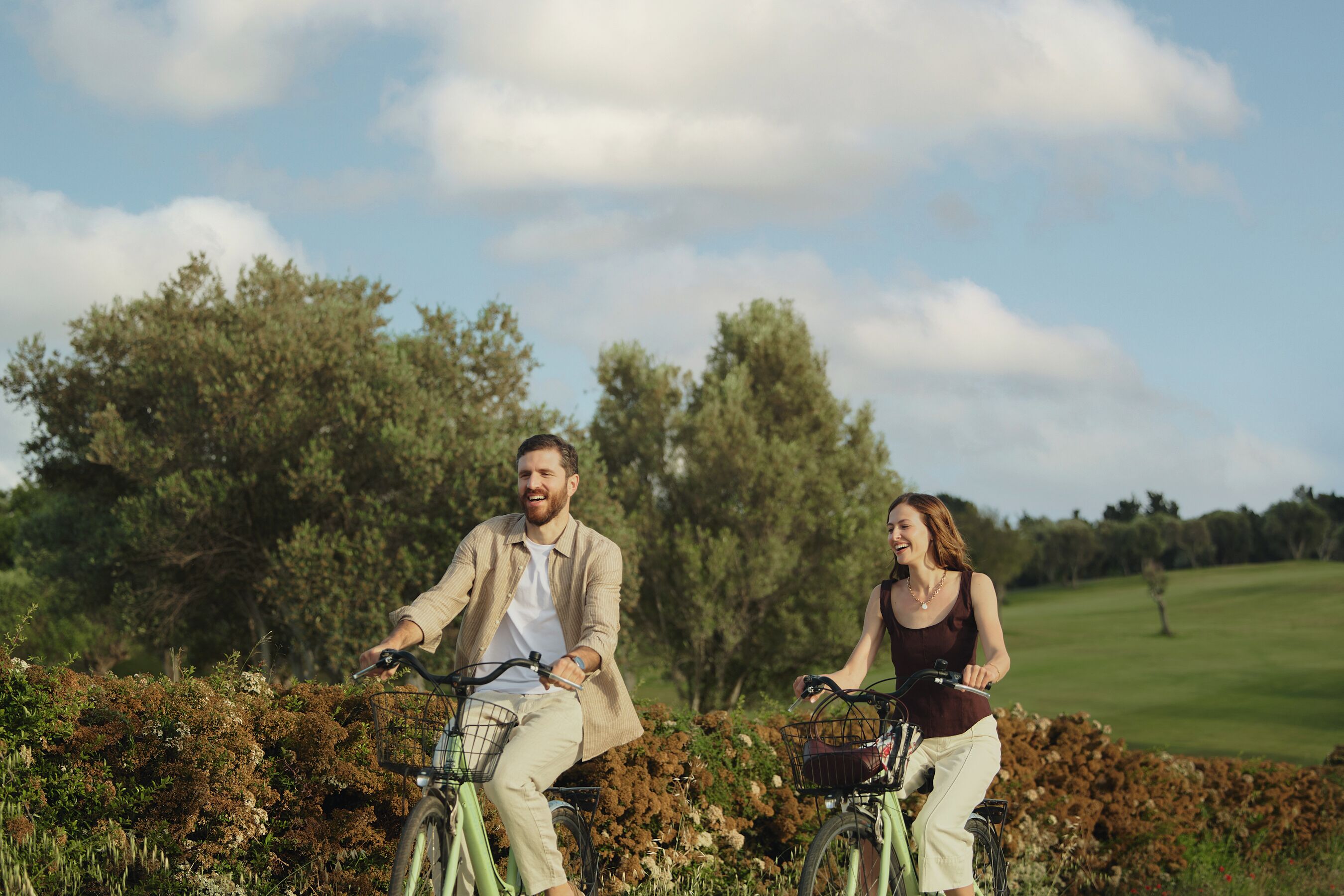 Duas pessoas de bicicleta em ambiente natural, com árvores e área gramada.
