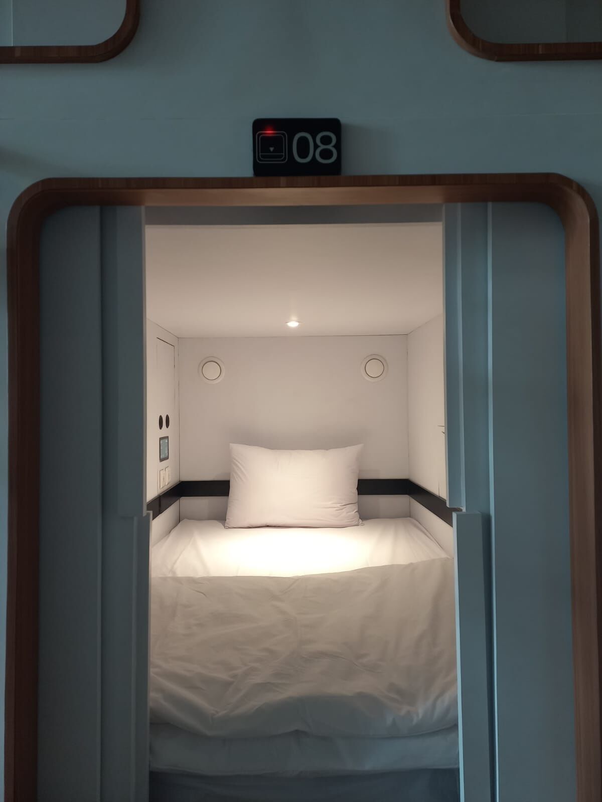 Quarto de hotel pequeno com uma cama de solteiro, uma mesa de cabeceira e um relógio na parede marcando 08:00.