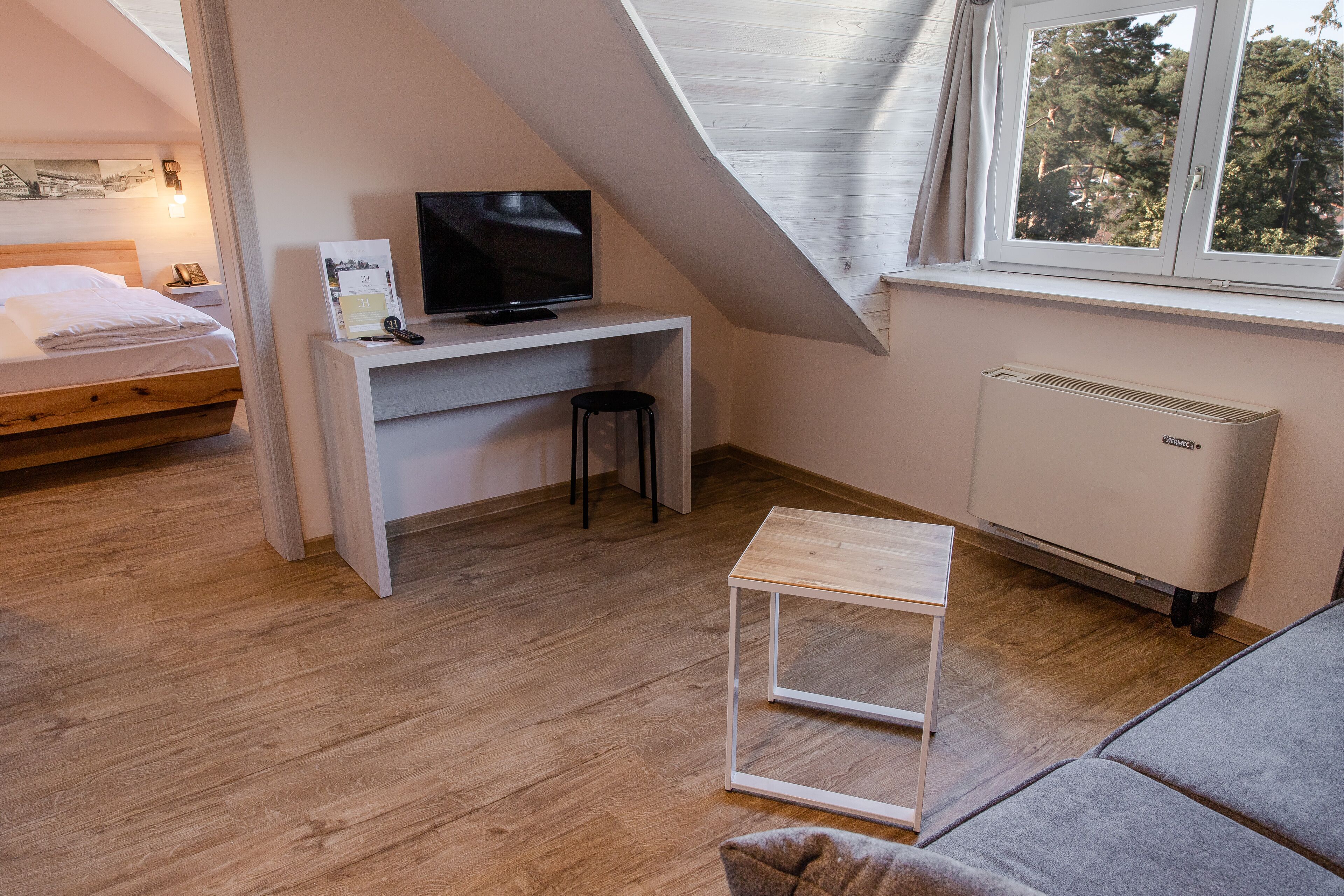 Quarto com cama, escrivaninha com televisão, radiador e janela com vista para árvores.