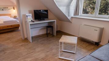 Quarto com cama, escrivaninha com televisão, radiador e janela com vista para árvores.
