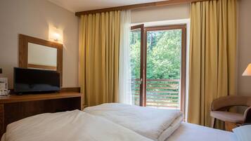 Quarto de hotel com cama, televisão sobre suporte de madeira, uma cadeira e janela com cortinas.