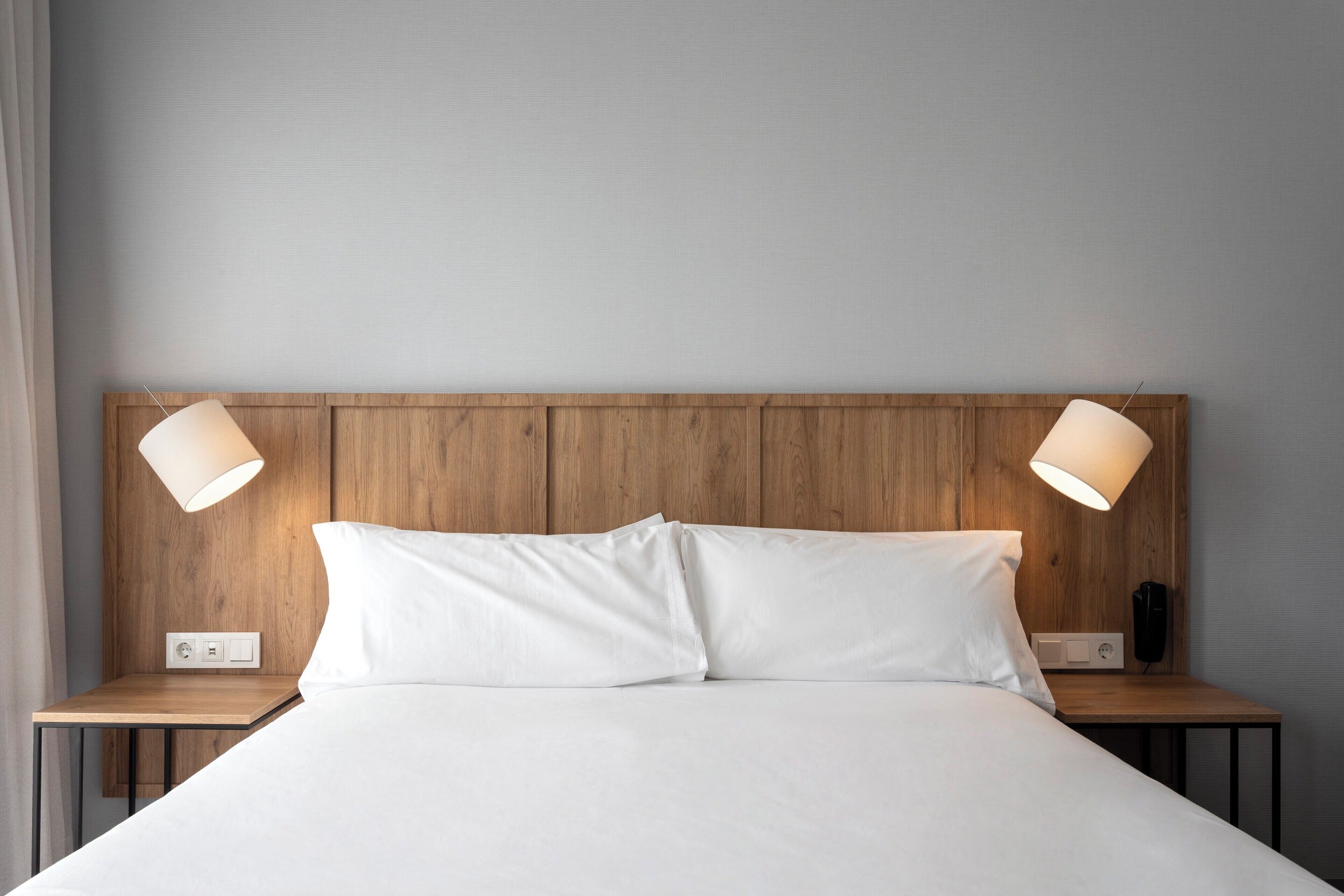 Cama com lençóis brancos, duas mesas de cabeceira com abajures e cabeceira de madeira.